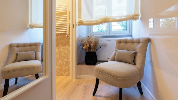 Um quarto com duas poltronas bege, um vaso preto com flores secas e uma janela com cortinas brancas.