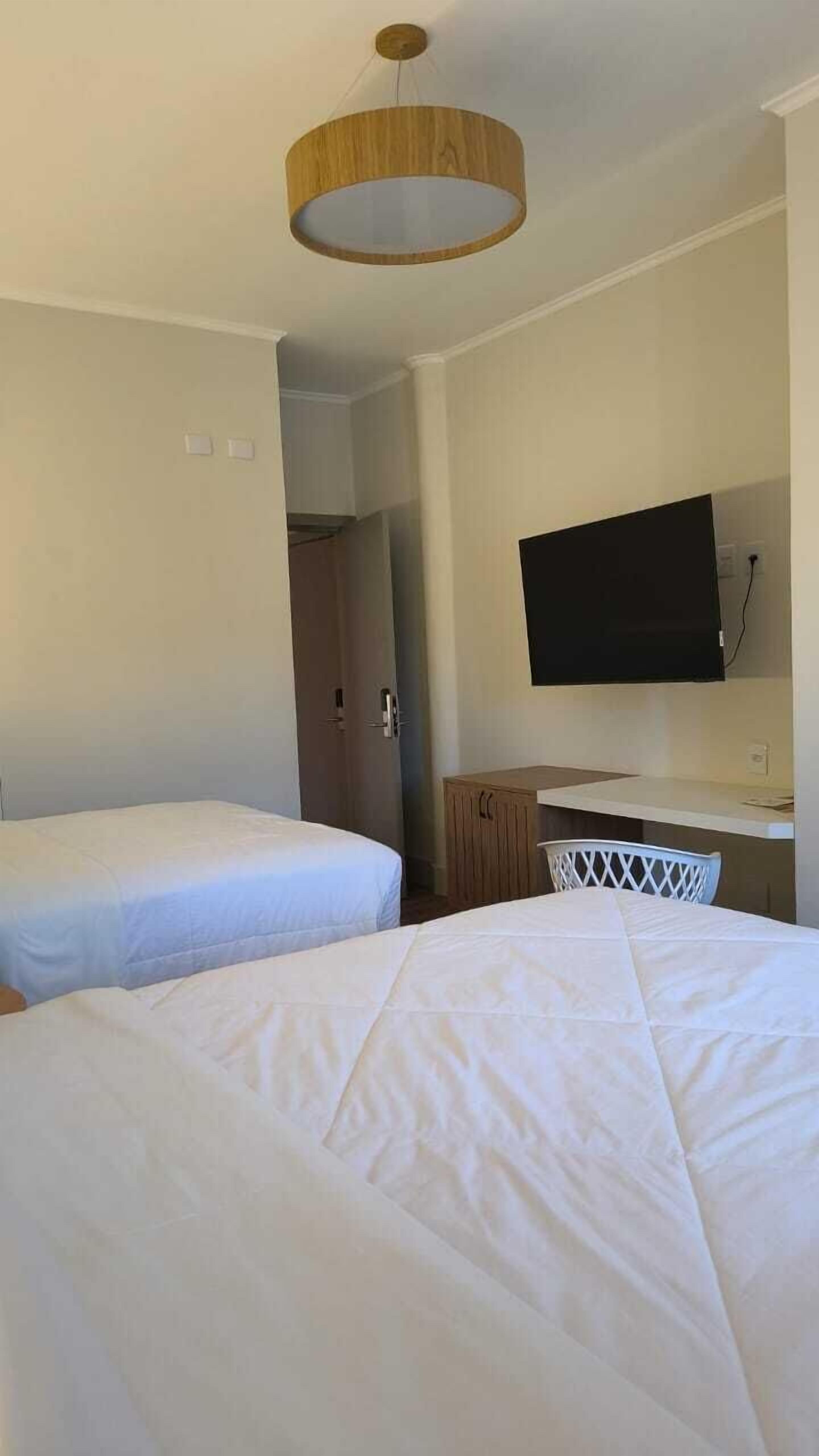 Quarto de hotel com duas camas, uma luminária de teto em madeira, uma televisão fixada na parede e uma pequena escrivaninha com cadeira.