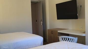 Quarto de hotel com duas camas, uma luminária de teto em madeira, uma televisão fixada na parede e uma pequena escrivaninha com cadeira.