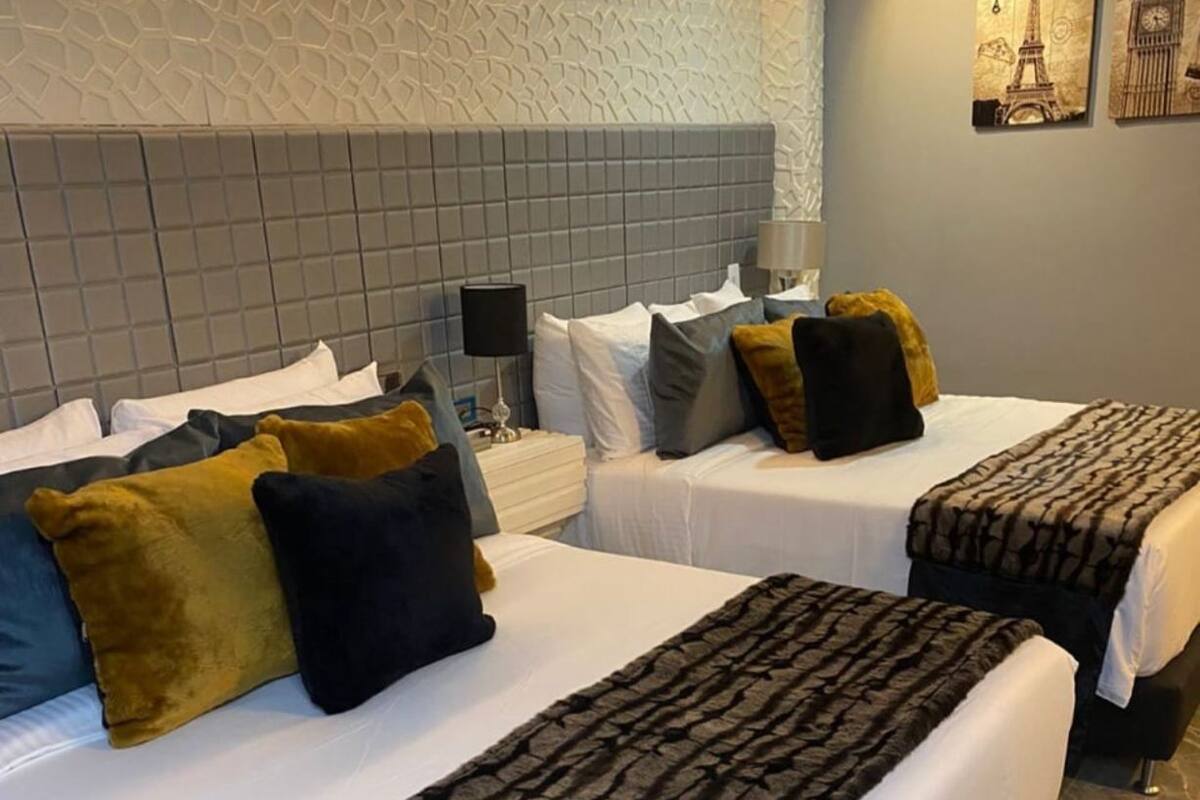 Quarto de hotel com duas camas, cada uma com uma colcha estampada e várias almofadas. O quarto possui uma parede azulejada e piso de mármore.