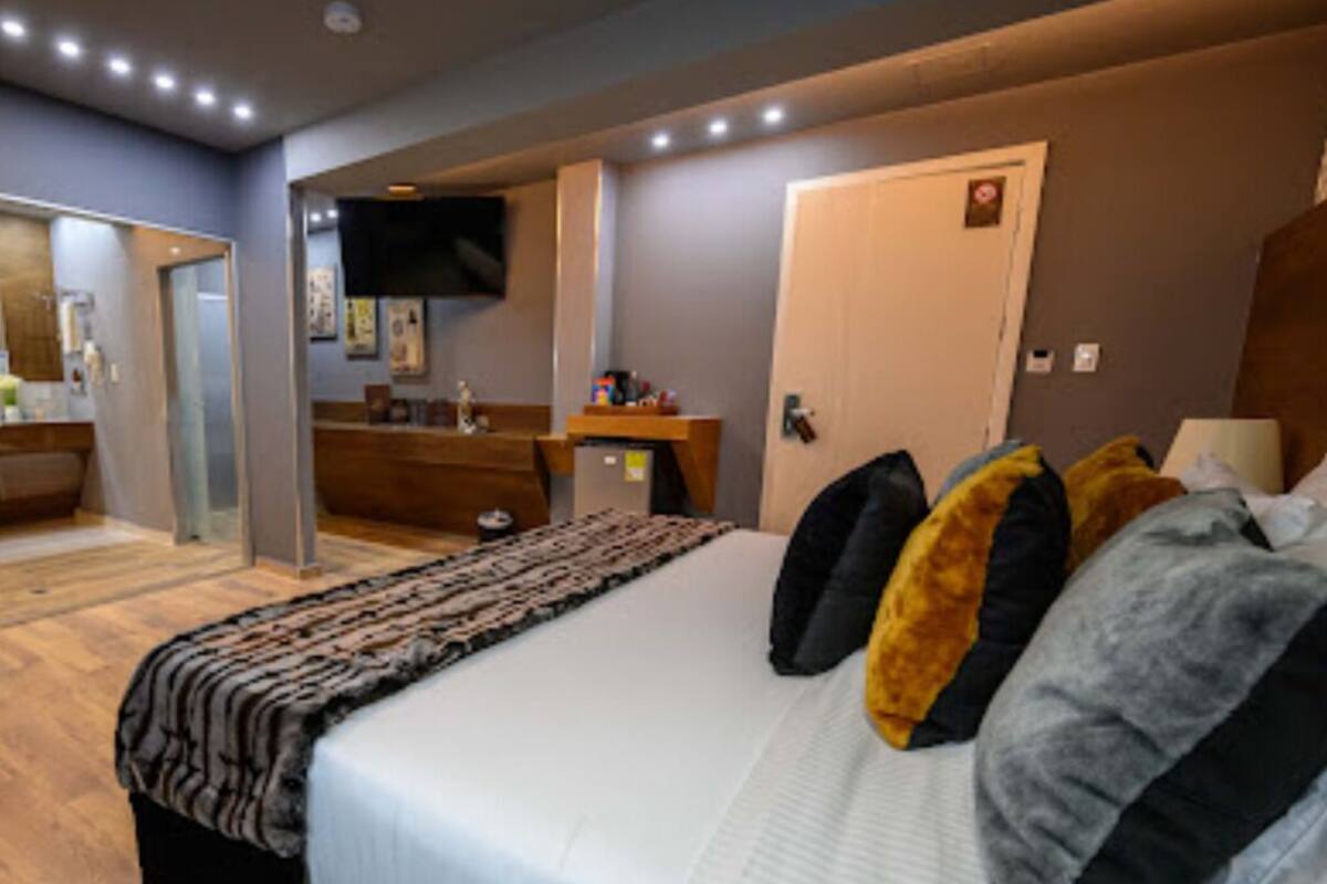 Quarto de hotel moderno com uma cama grande, televisão de tela plana na parede, escrivaninha de madeira e banheiro visível através de uma porta aberta.