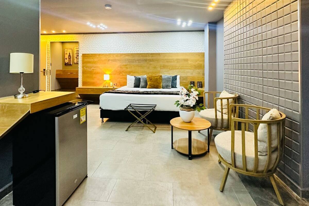 Quarto de hotel moderno com cama, pequena cozinha, área de estar com cadeiras e um detalhe decorativo na parede.
