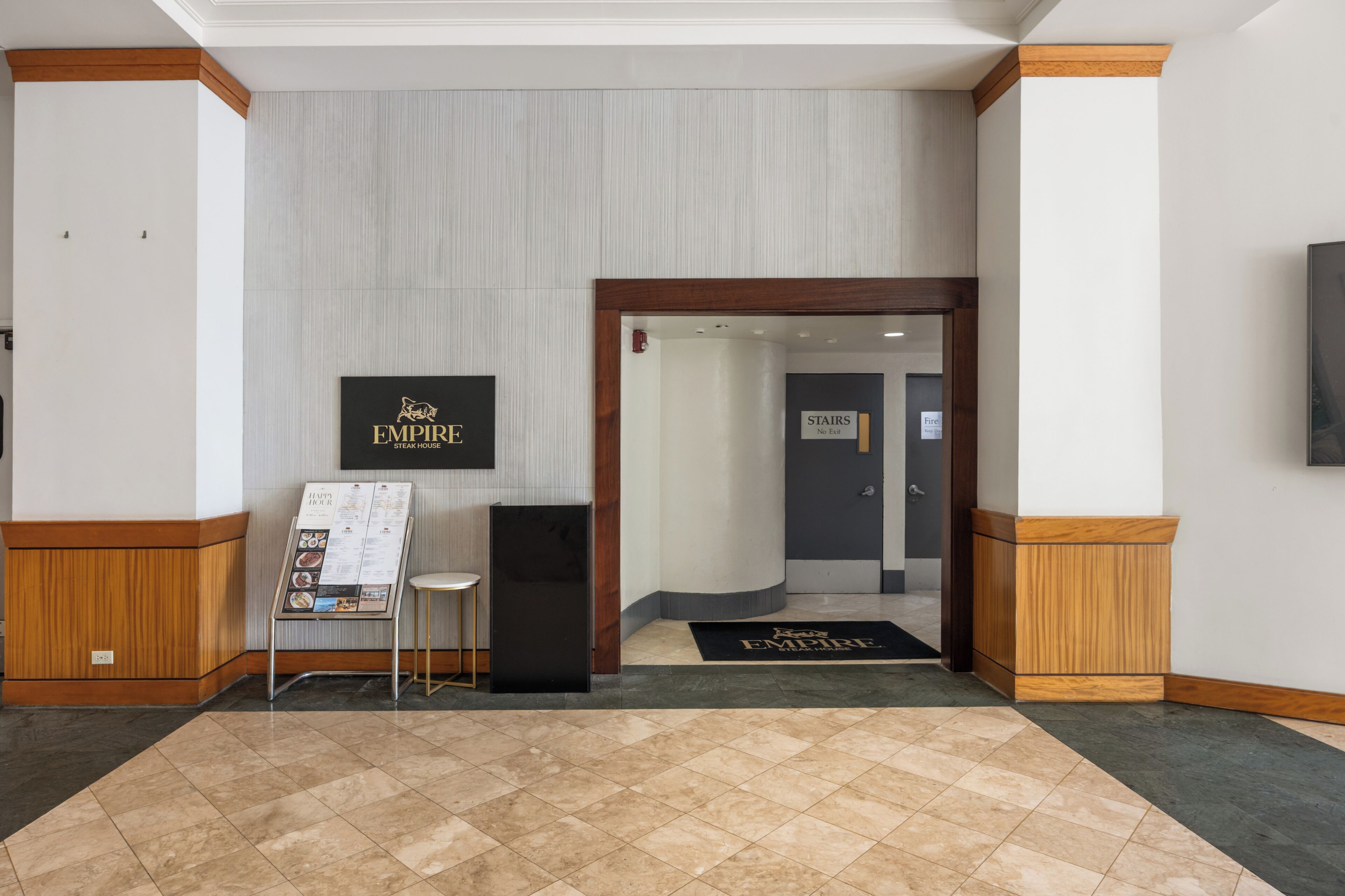 Um saguão de hotel com uma mesa de recepção, uma placa preta com o texto 'EMPIRE STEAK HOUSE' e um aviso de 'HAPPY HOUR'.