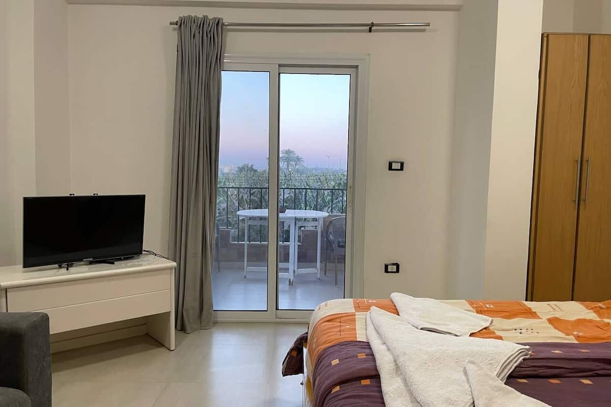 Quarto de hotel com cama, televisão sobre uma mesa de cabeceira, guarda-roupa e porta de vidro deslizante que dá para área externa com mesa e cadeiras.