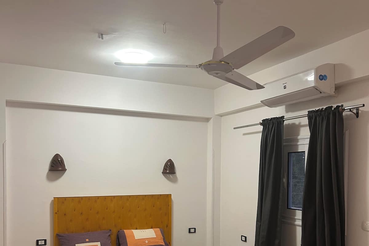 Quarto com cama de madeira, ventoinha no teto, ar condicionado e janela com cortinas.