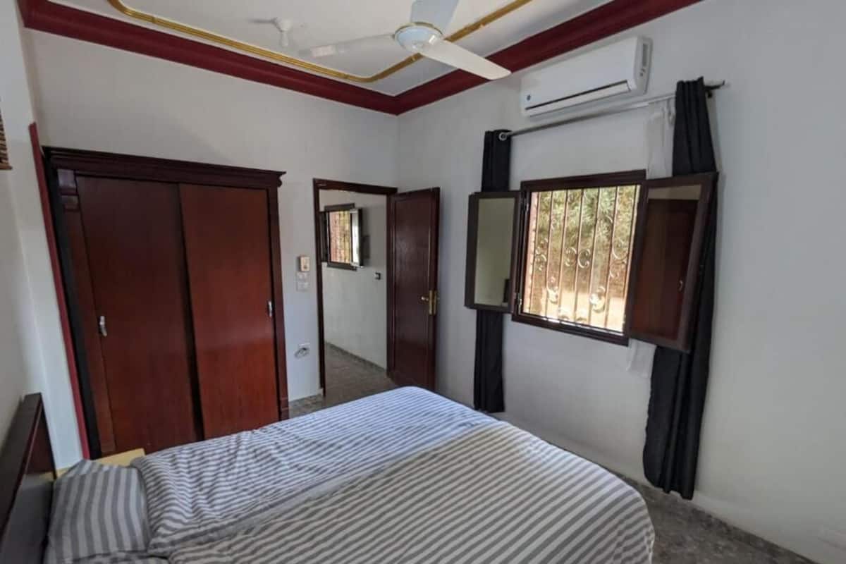 Quarto com cama, ventilador de teto, ar condicionado, janela com grades e porta.