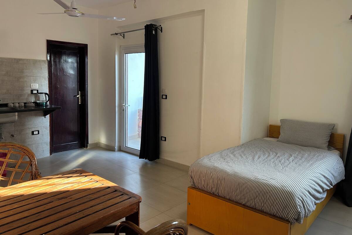 Quarto com cama, mesa de madeira, cadeira, ventoinha no teto e vista para área externa através de uma porta.