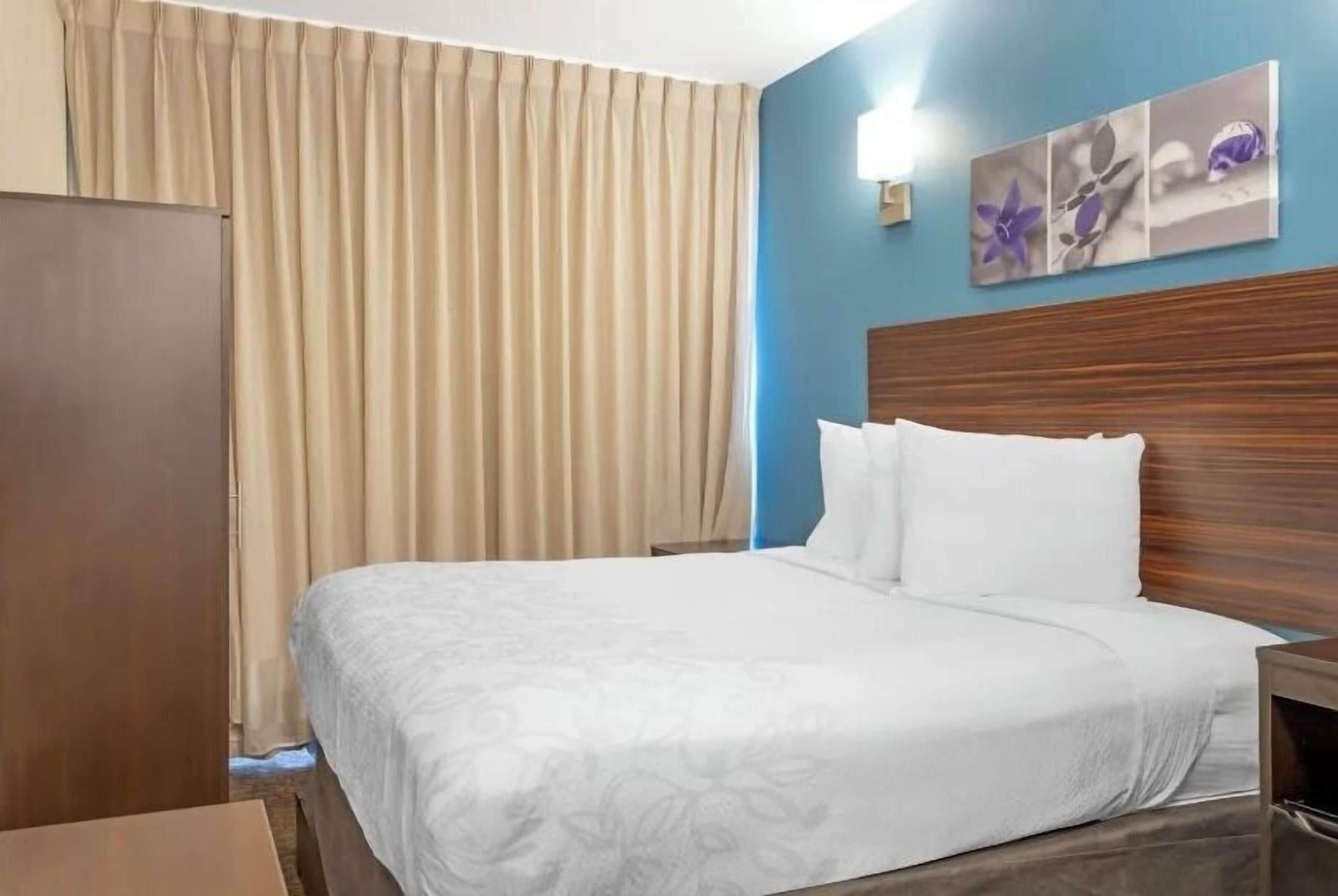 Quarto de hotel com cama, cabeceira de madeira, cortinas bege e parede com pintura floral.