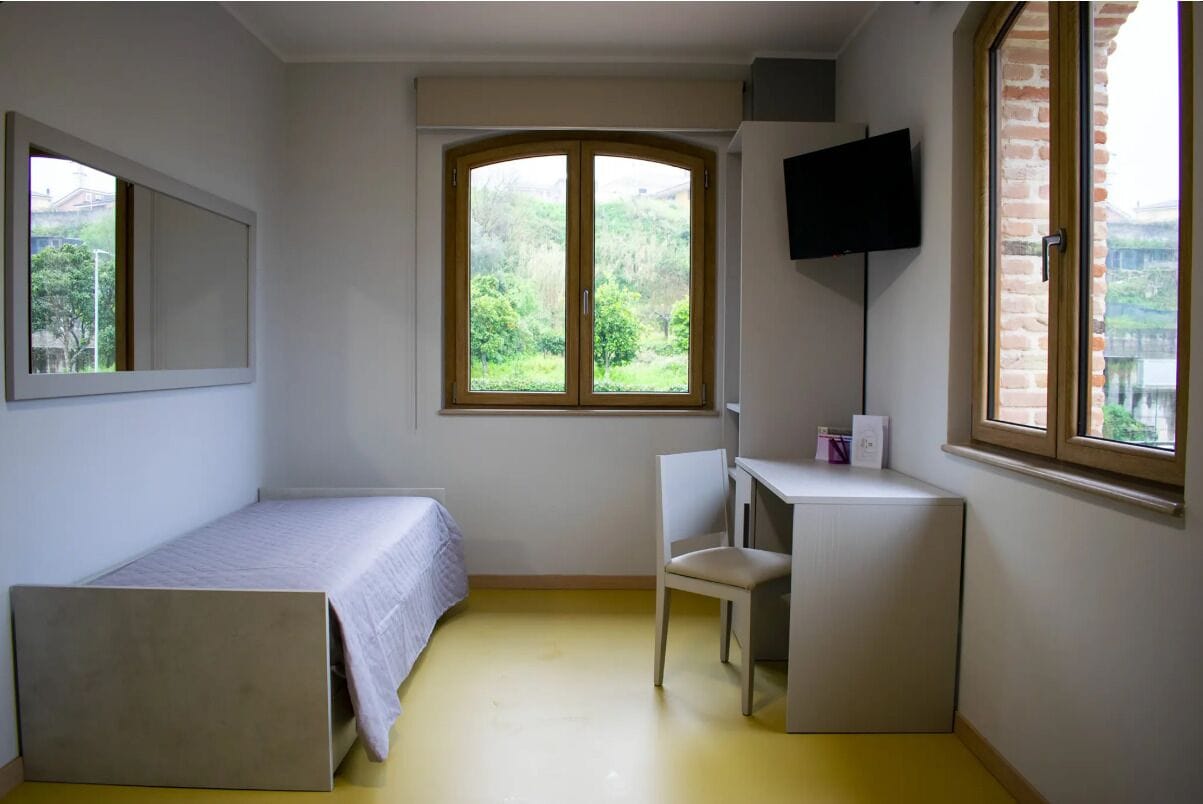 Quarto pequeno e individual com cama, escrivaninha, cadeira, televisão e janela com vista para o verde.