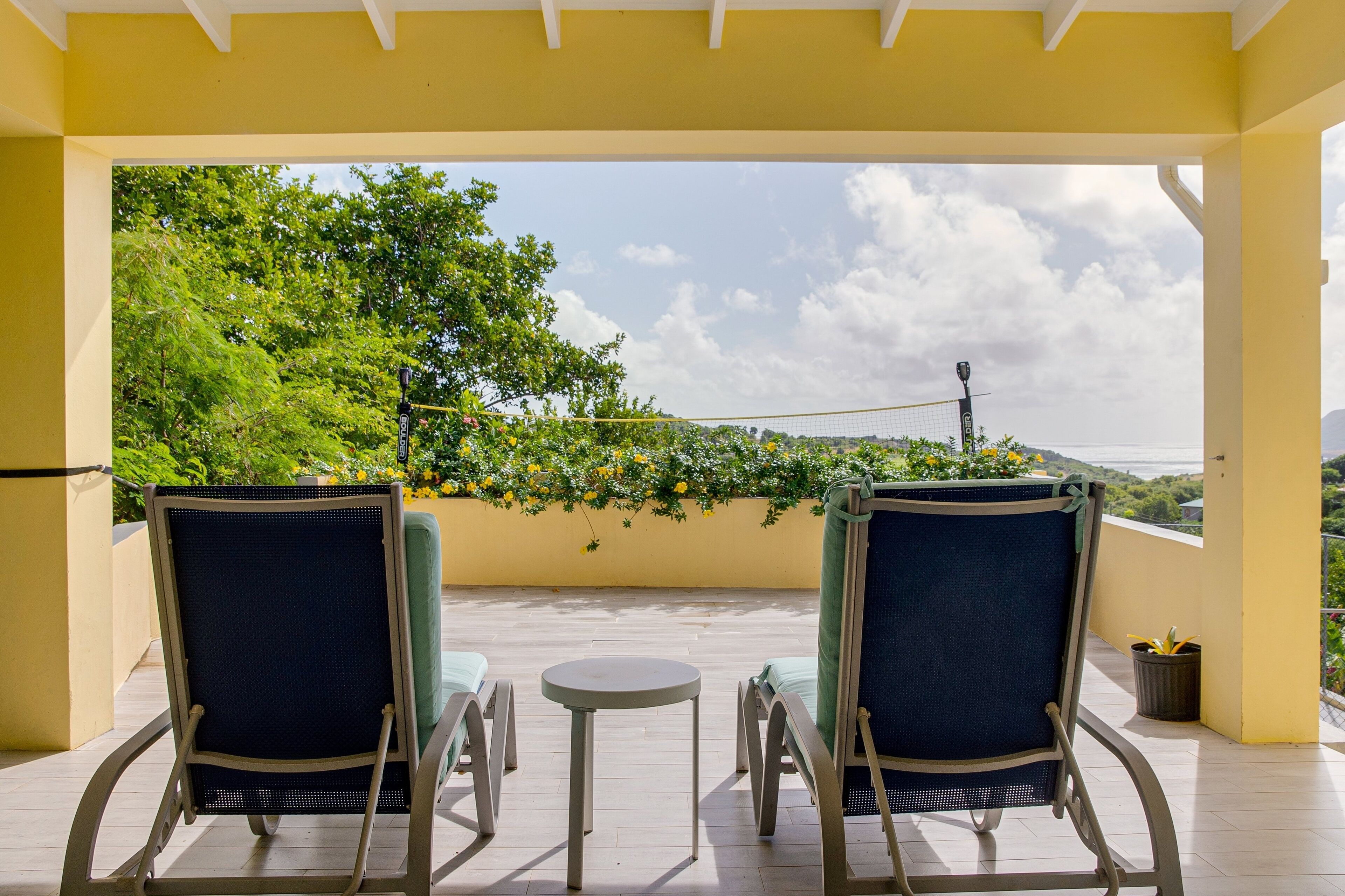 Varanda com duas cadeiras e uma pequena mesa, com vista para um jardim e o mar.