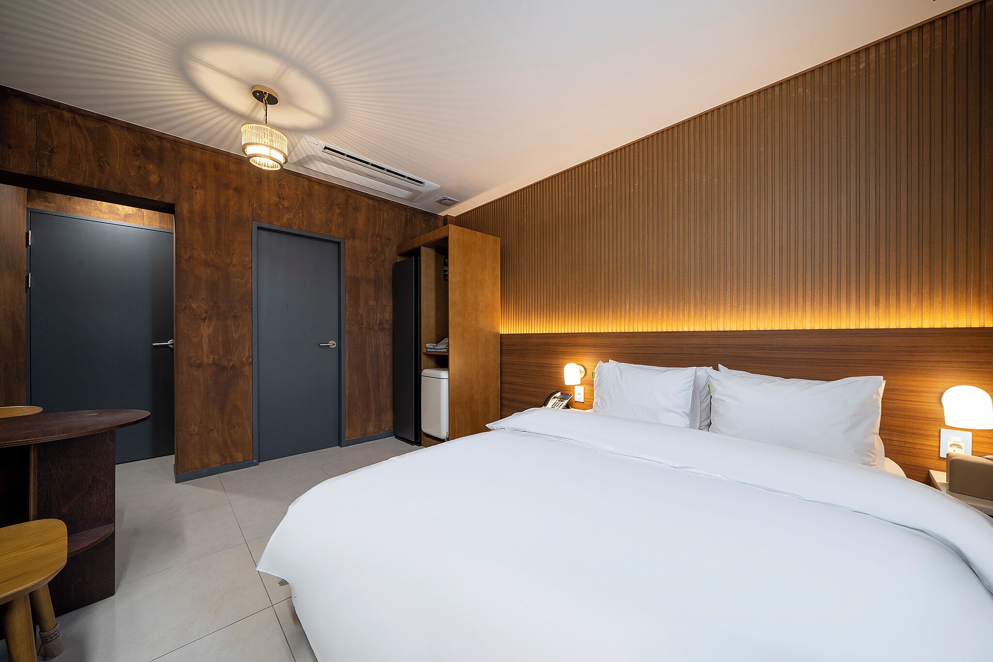 Quarto de hotel com uma cama grande, uma escrivaninha e uma cadeira. Possui cabeceira de madeira e uma luminária fixada na parede.