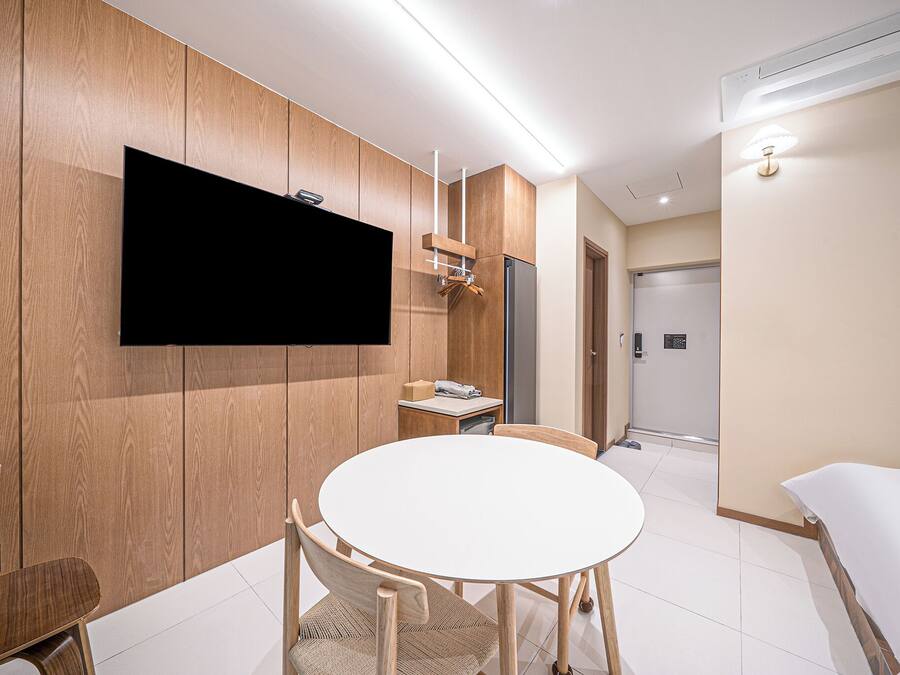 Quarto de hotel moderno com mesa de jantar redonda, paredes revestidas de madeira, TV de tela plana na parede, pequena área de cozinha e espaço de dormir separado.