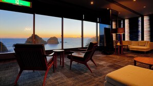 Quarto de hotel moderno com grandes janelas que oferecem vista para o mar e falésias ao pôr do sol. Há duas cadeiras de madeira, um sofá e uma pequena mesa.