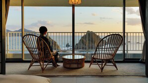 Quarto com uma grande janela com vista para a costa, duas cadeiras de vime, uma mesa redonda e um abajur.