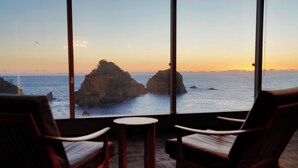Quarto com uma grande janela com vista para a paisagem costeira, com formações rochosas e o mar. Há duas cadeiras de madeira e uma pequena mesa redonda no quarto.