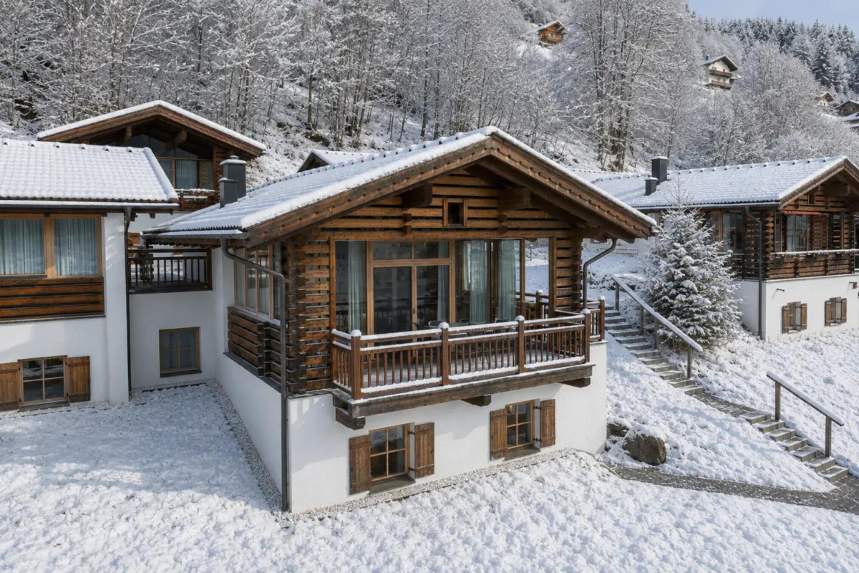 Um chalé de madeira com varanda, cercado por árvores cobertas de neve e um cenário montanhoso.