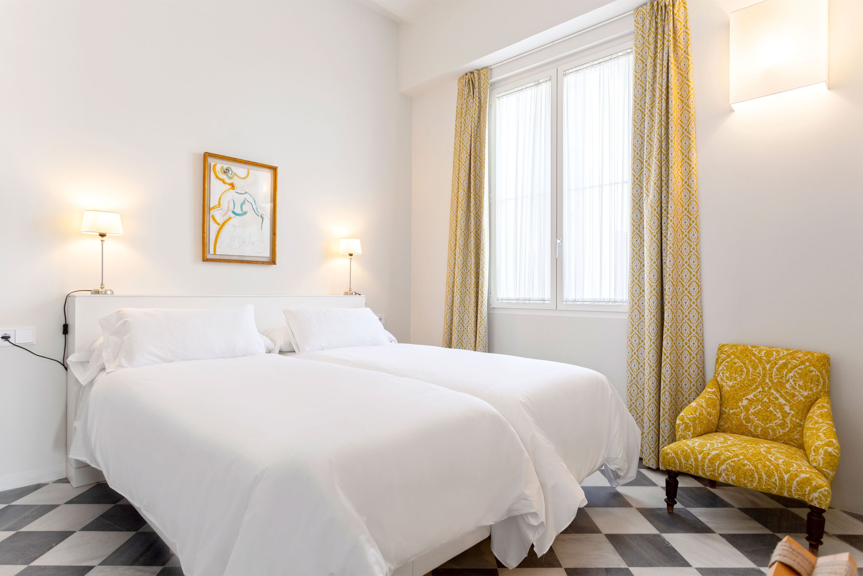 Quarto de hotel com uma cama grande, duas luminárias de cabeceira, uma obra de arte emoldurada, uma janela com cortinas e uma poltrona estampada.
