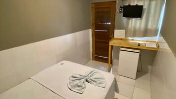 Quarto de hotel pequeno com uma cama de solteiro, uma escrivaninha com televisão e um aparelho de ar condicionado.