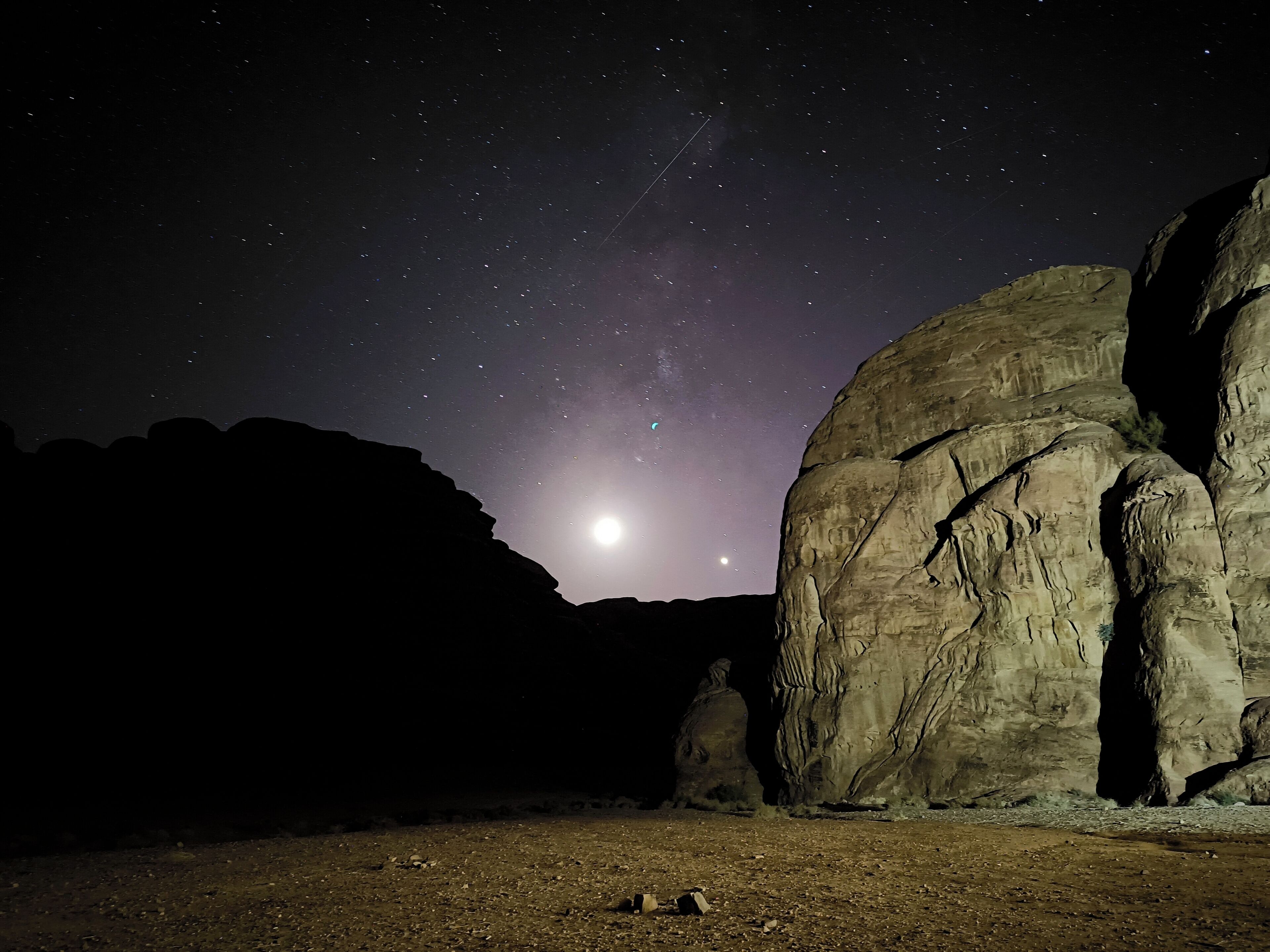 Céu noturno com estrelas e a lua, paisagem rochosa e uma grande formação rochosa.