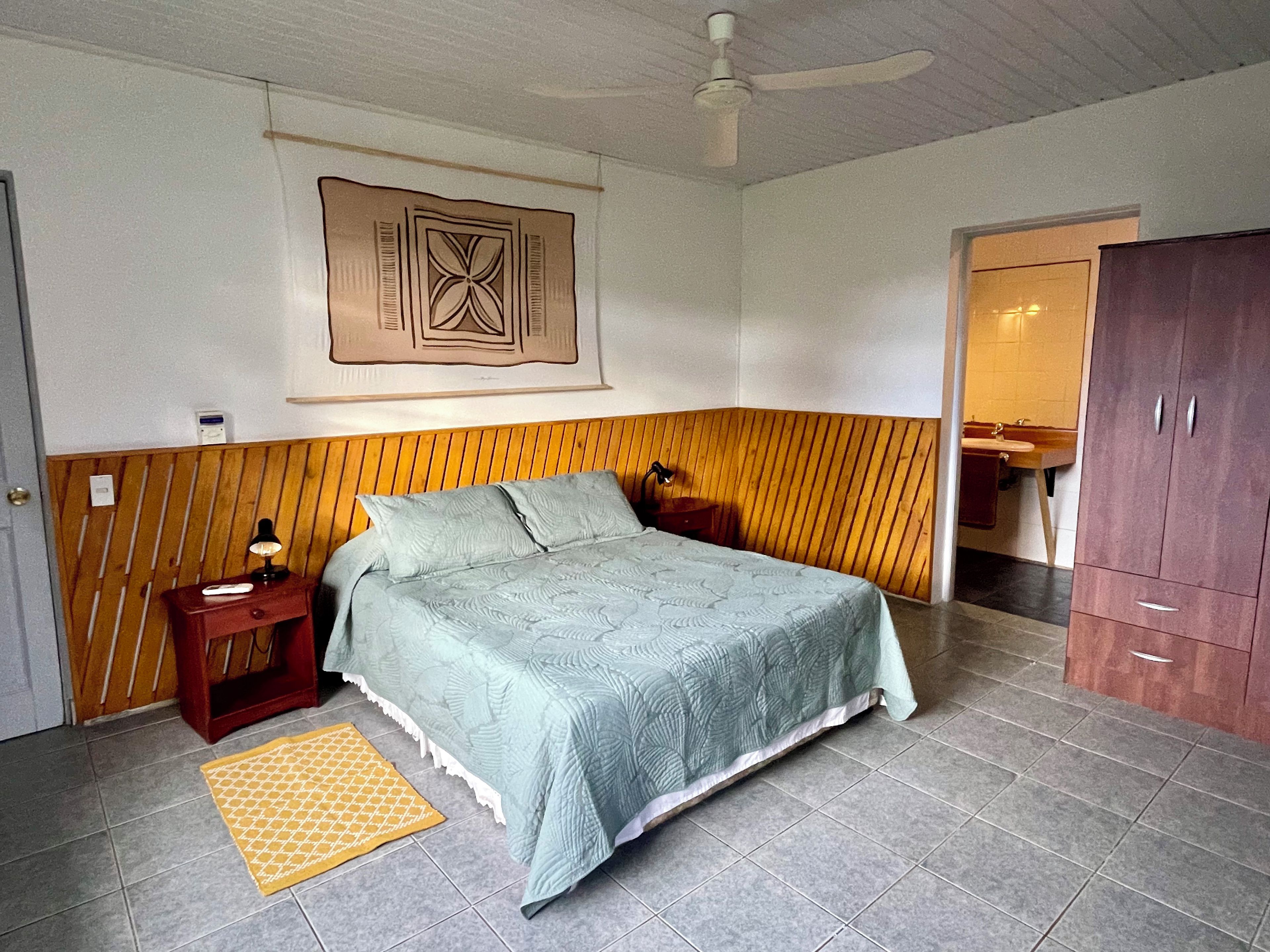 Quarto com cama, mesa de cabeceira, luminária, obra de arte na parede, ventoinha no teto e banheiro visível por uma porta aberta.