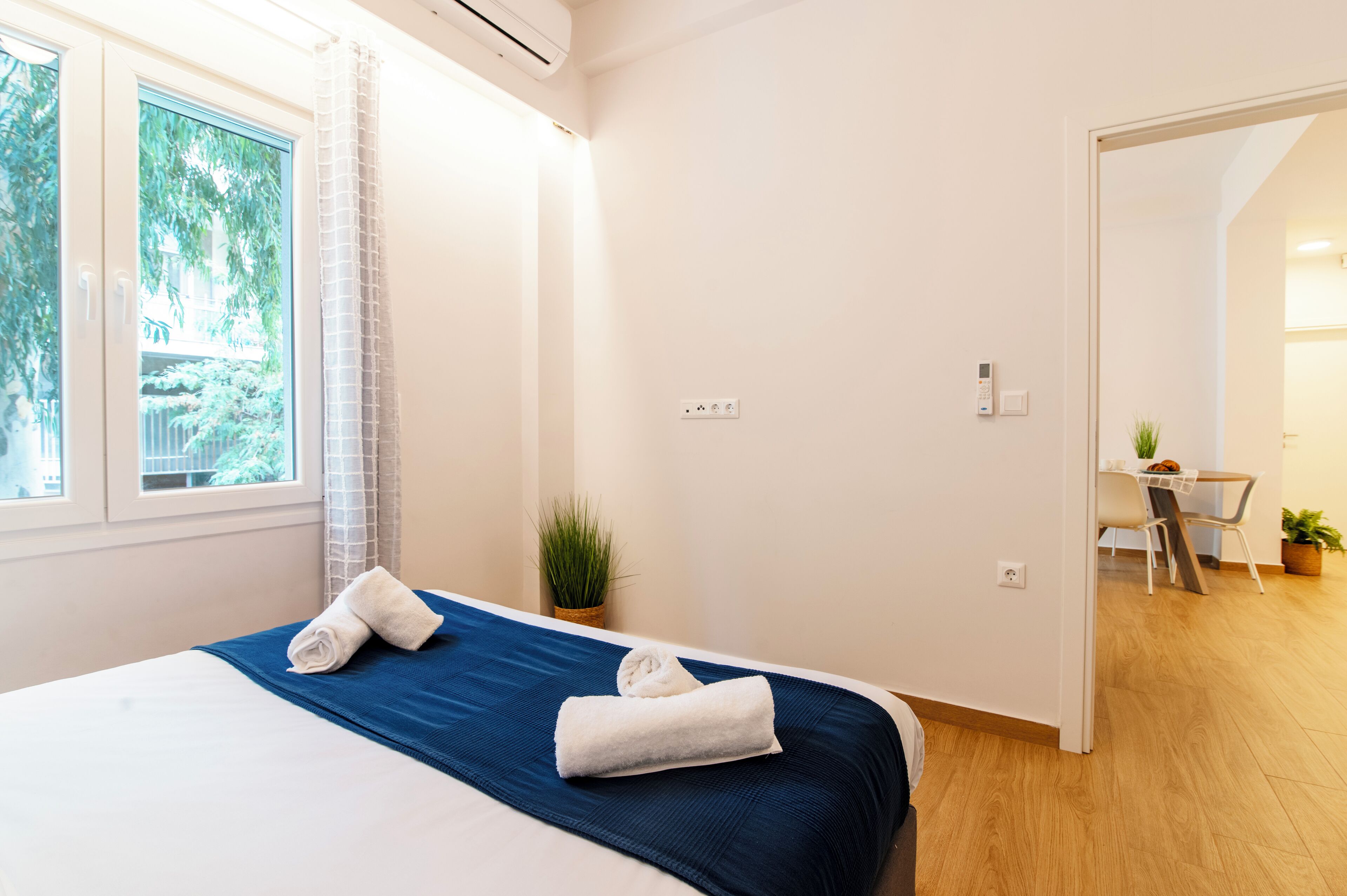Quarto de hotel com cama, uma pequena mesa com cadeiras, um vaso de planta e vista para o verde lá fora.