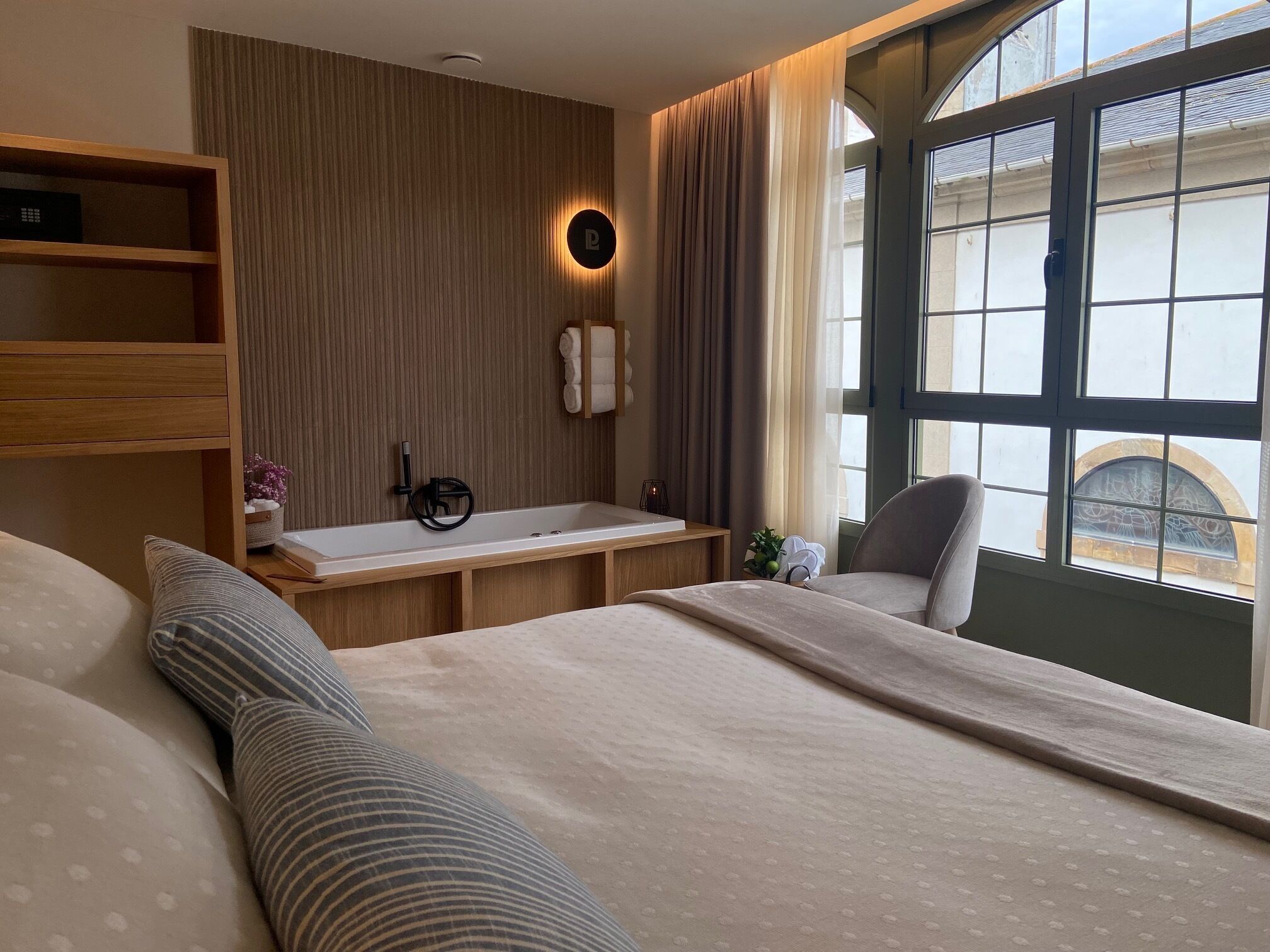 Quarto de hotel com uma cama grande, mesa de cabeceira, lavatório embutido e janela com vista para a cidade.