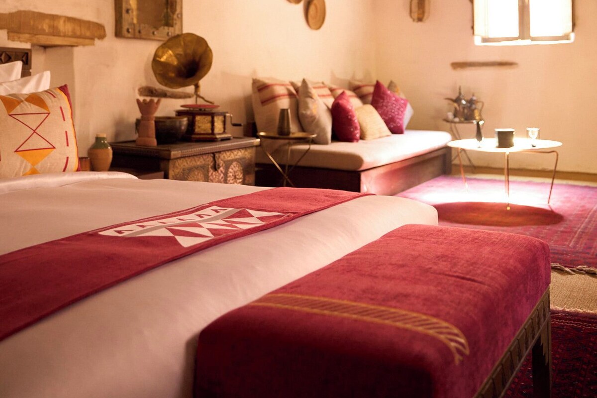 Quarto tradicional com teto de madeira, uma cama com manta vermelha, uma mesinha com abajur e uma janela com luz natural.
