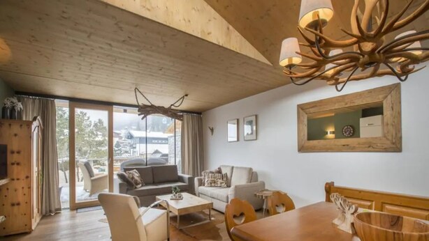 Uma sala de estar moderna com teto de madeira, uma mesa de jantar com cadeiras, um sofá e uma janela ampla com vista para uma paisagem coberta de neve.