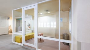 Quarto com porta de vidro deslizante, um banco amarelo, uma mesa pequena e uma lata de lixo.