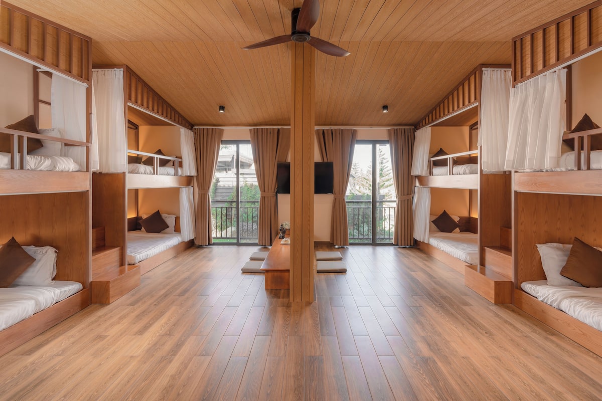 Quarto com beliches, piso de madeira, ventilador de teto e grandes janelas.
