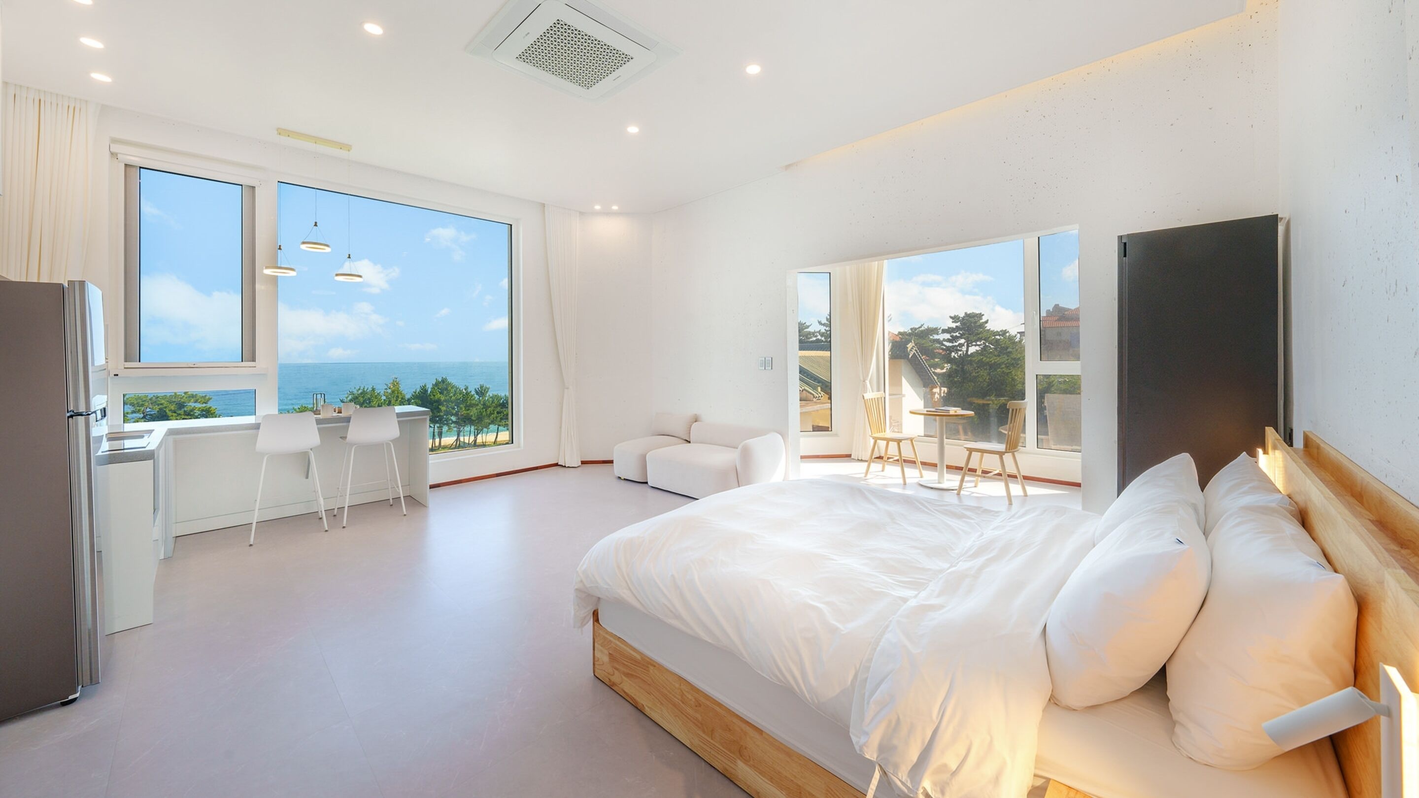 Quarto moderno com uma cama grande, uma escrivaninha e cadeiras, amplas janelas com vista para o mar e uma varanda com mesa e cadeiras.