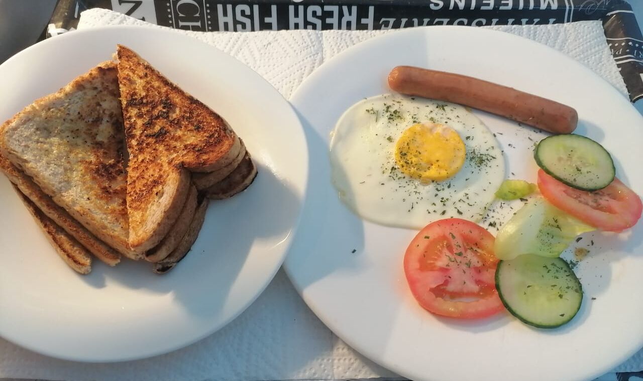 Um prato com torrada, ovo frito, salsicha e rodelas de tomate e pepino.