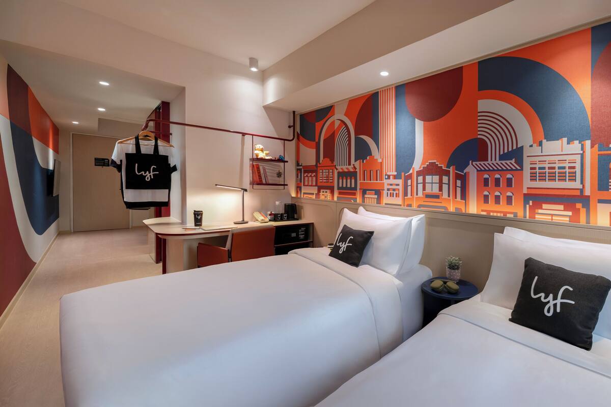 Quarto de hotel com duas camas, uma escrivaninha, uma cadeira, uma pequena mesa com uma planta e um mural na parede.
