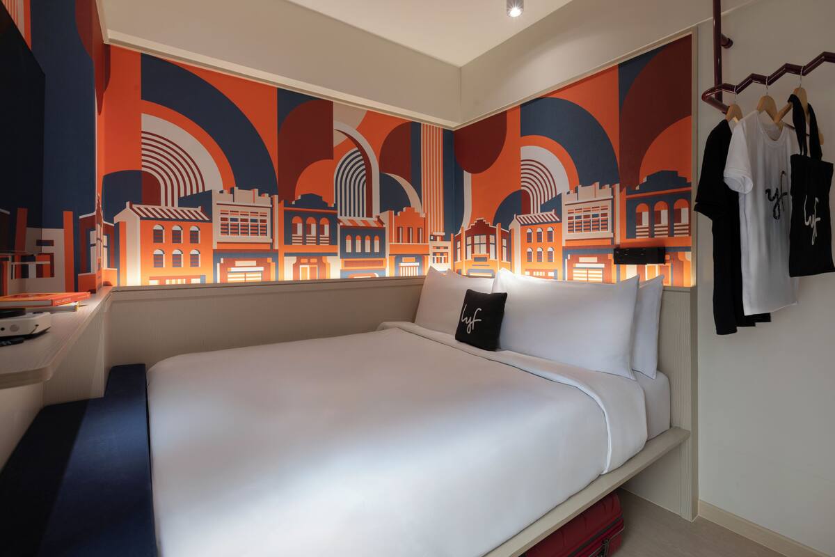 Quarto de hotel com cama, mesa de cabeceira, um mural na parede e um cabideiro.