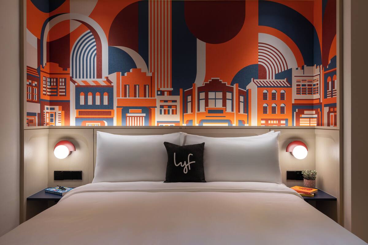 Quarto de hotel com uma cama, um travesseiro com a inscrição 'lyf', duas mesas de cabeceira, uma obra de arte na parede e uma luz noturna.
