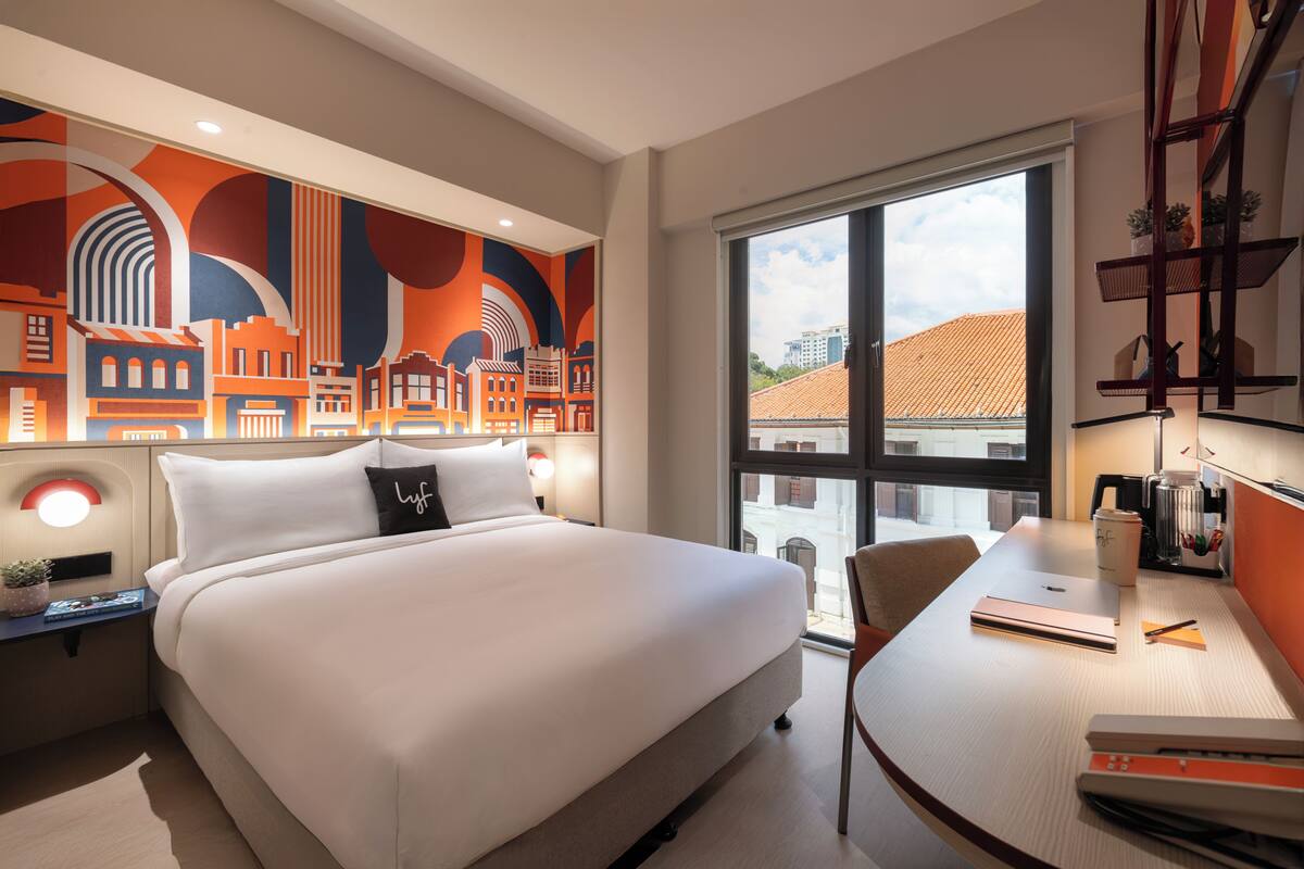 Quarto de hotel moderno com uma cama grande, uma escrivaninha com cadeira, uma janela ampla e um mural com vista urbana na parede.