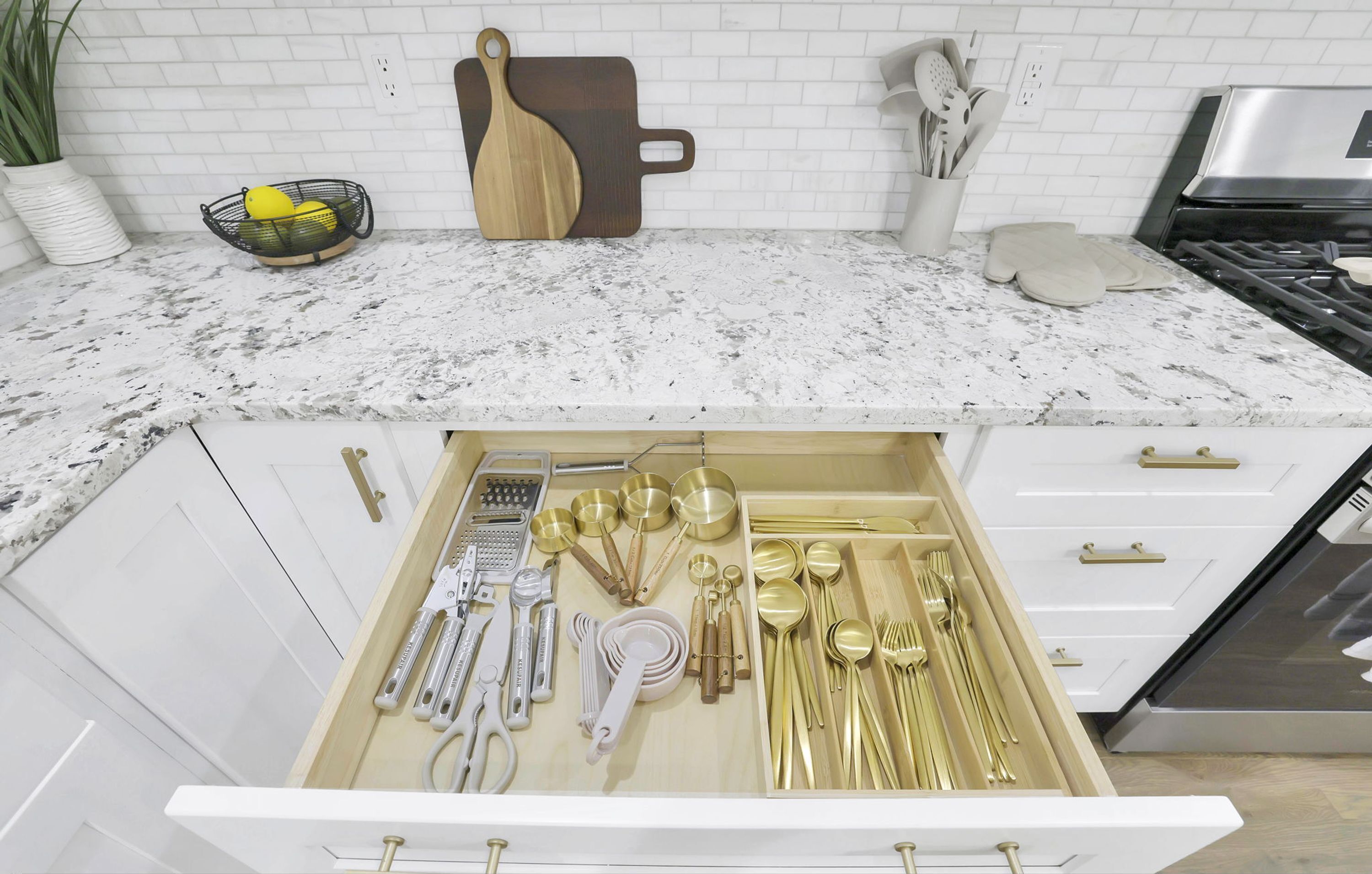 Uma cozinha com bancada de mármore, tábua de corte de madeira e uma gaveta com utensílios de cozinha.