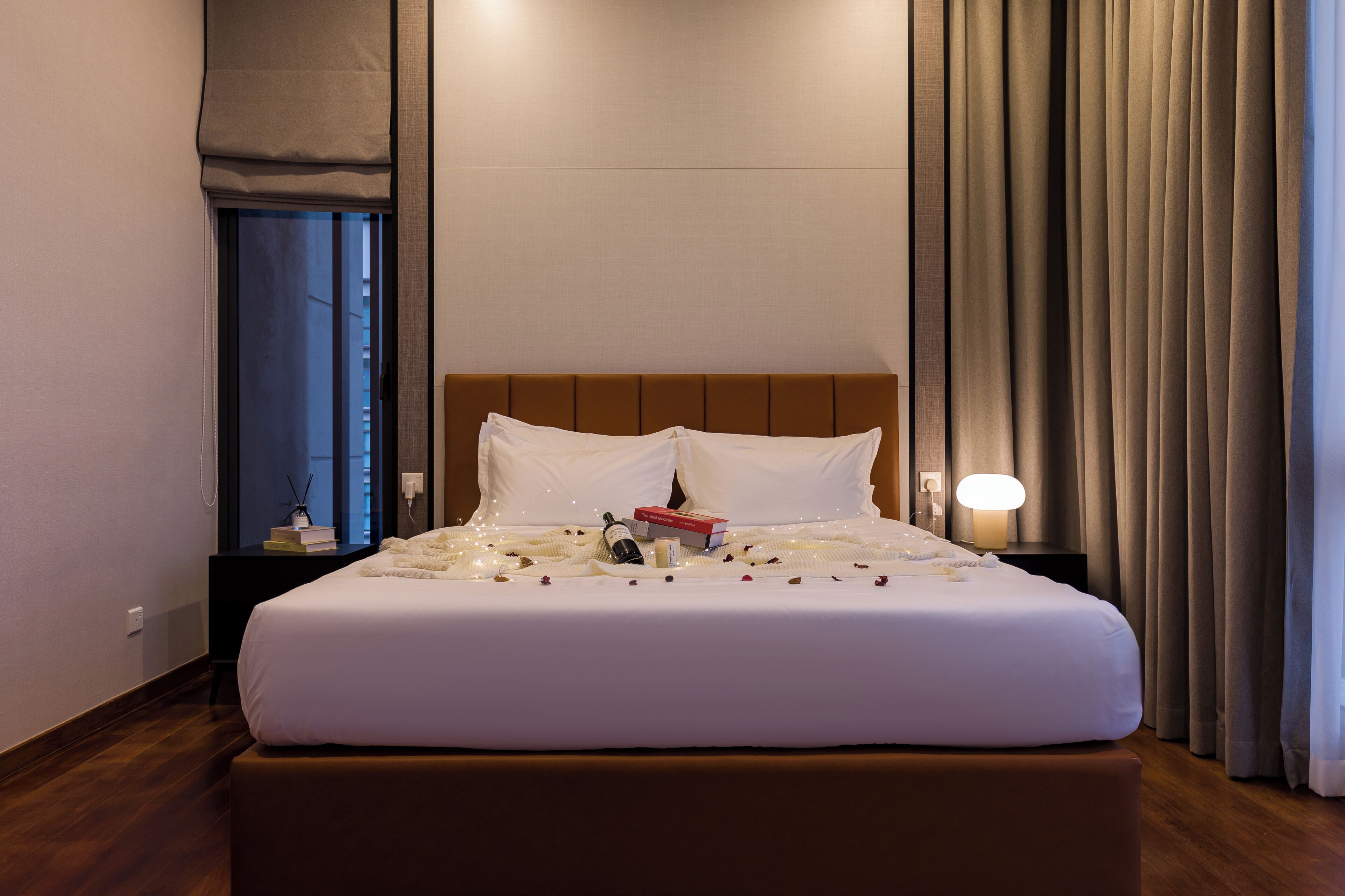 Uma cama bem arrumada com lençóis brancos, uma mesinha de cabeceira com abajur e uma janela com cortinas.