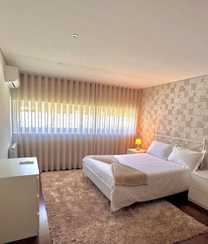 Quarto com cama, mesa de cabeceira, luminária, radiador e janela com cortinas.