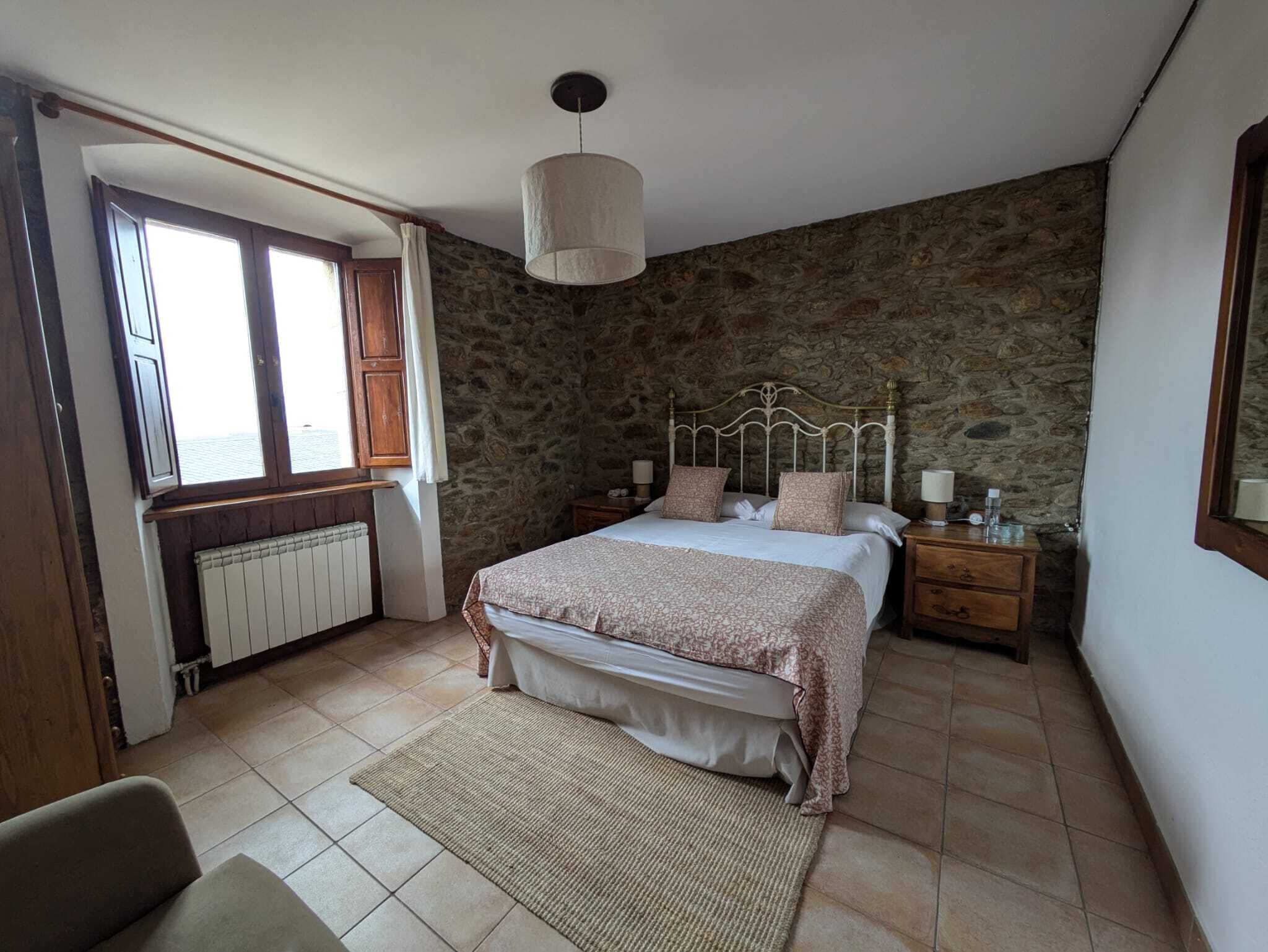 Um quarto com parede de pedra, uma cama com colcha estampada, uma mesa de cabeceira com abajur e uma janela com cortinas.