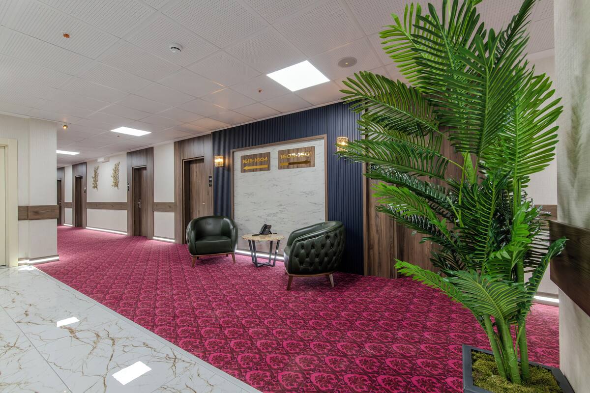 Um saguão de hotel com tapete vermelho, poltronas de couro, um balcão de recepção e uma grande planta em vaso.