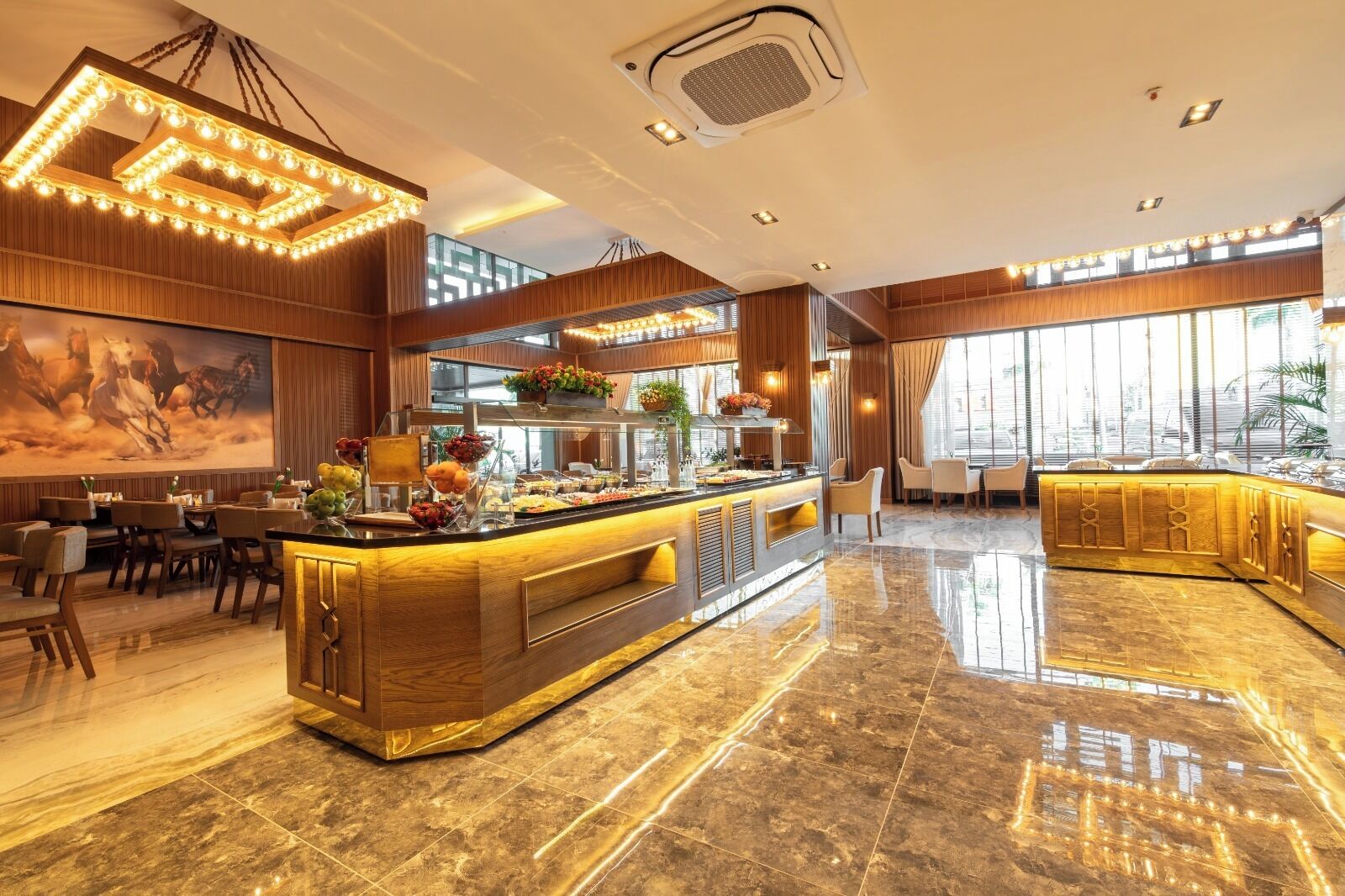 Um amplo saguão de hotel com área de buffet central, painéis de madeira e grandes janelas que oferecem iluminação natural.