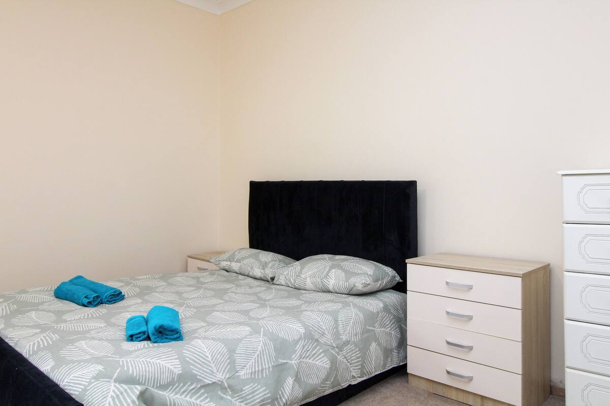 Uma cama bem arrumada com colcha e almofadas estampadas com folhas, um penteou branco com gavetas e uma cabeceira preta.
