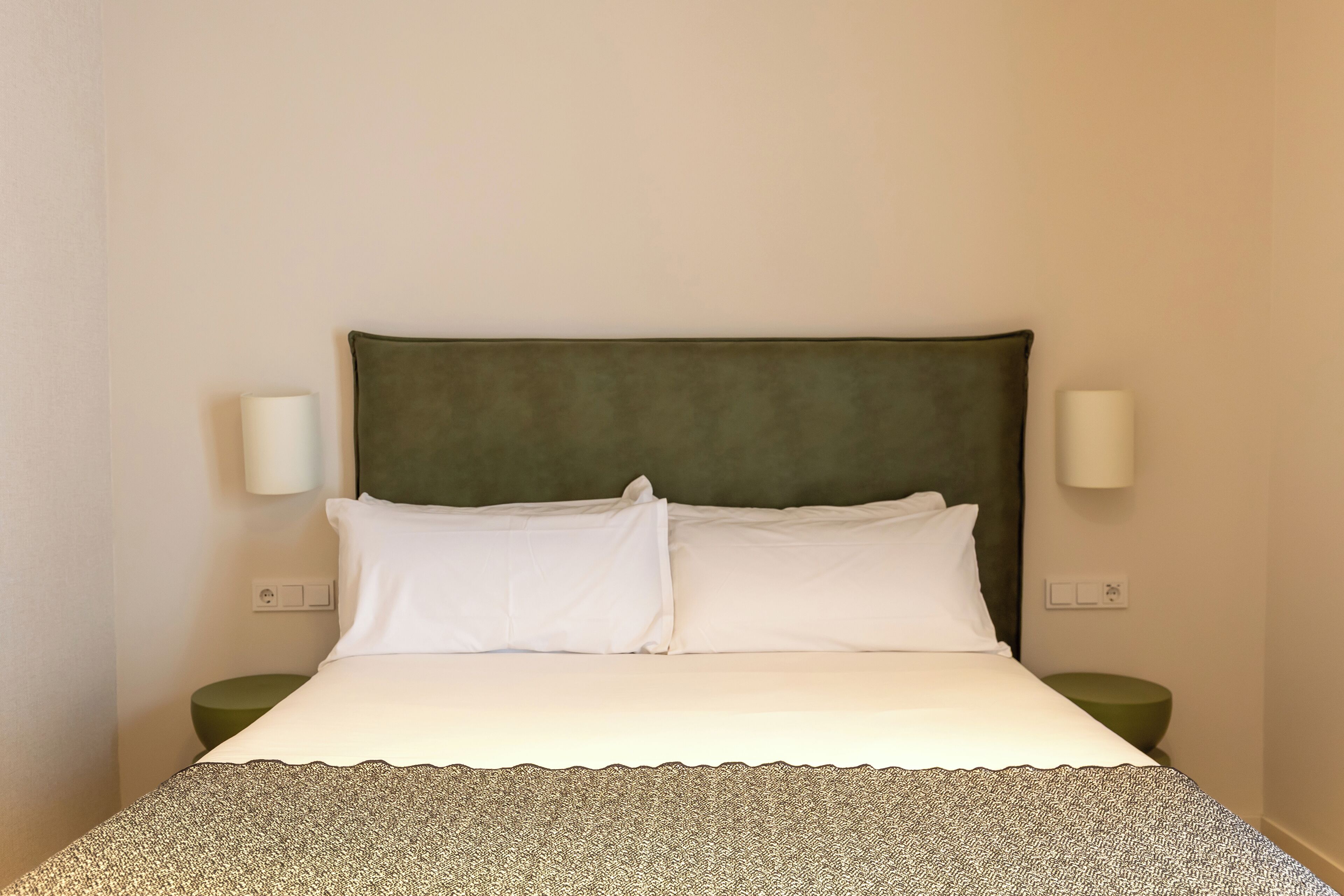Uma cama bem arrumada com cabeceira verde, almofadas brancas e colcha estampada. Há duas mesas de cabeceira verdes com abajures brancos.