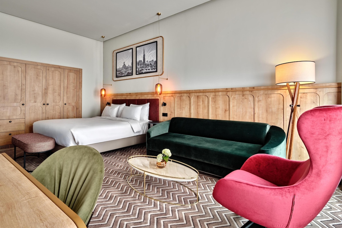 Quarto de hotel com cama, sofá verde, poltrona vermelha, guarda-roupa de madeira e luminária de chão.