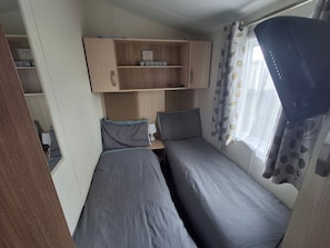 Quarto compacto com duas camas de solteiro, um armário de madeira com espaço para guardar, uma mesinha com abajur e uma janela com cortina.