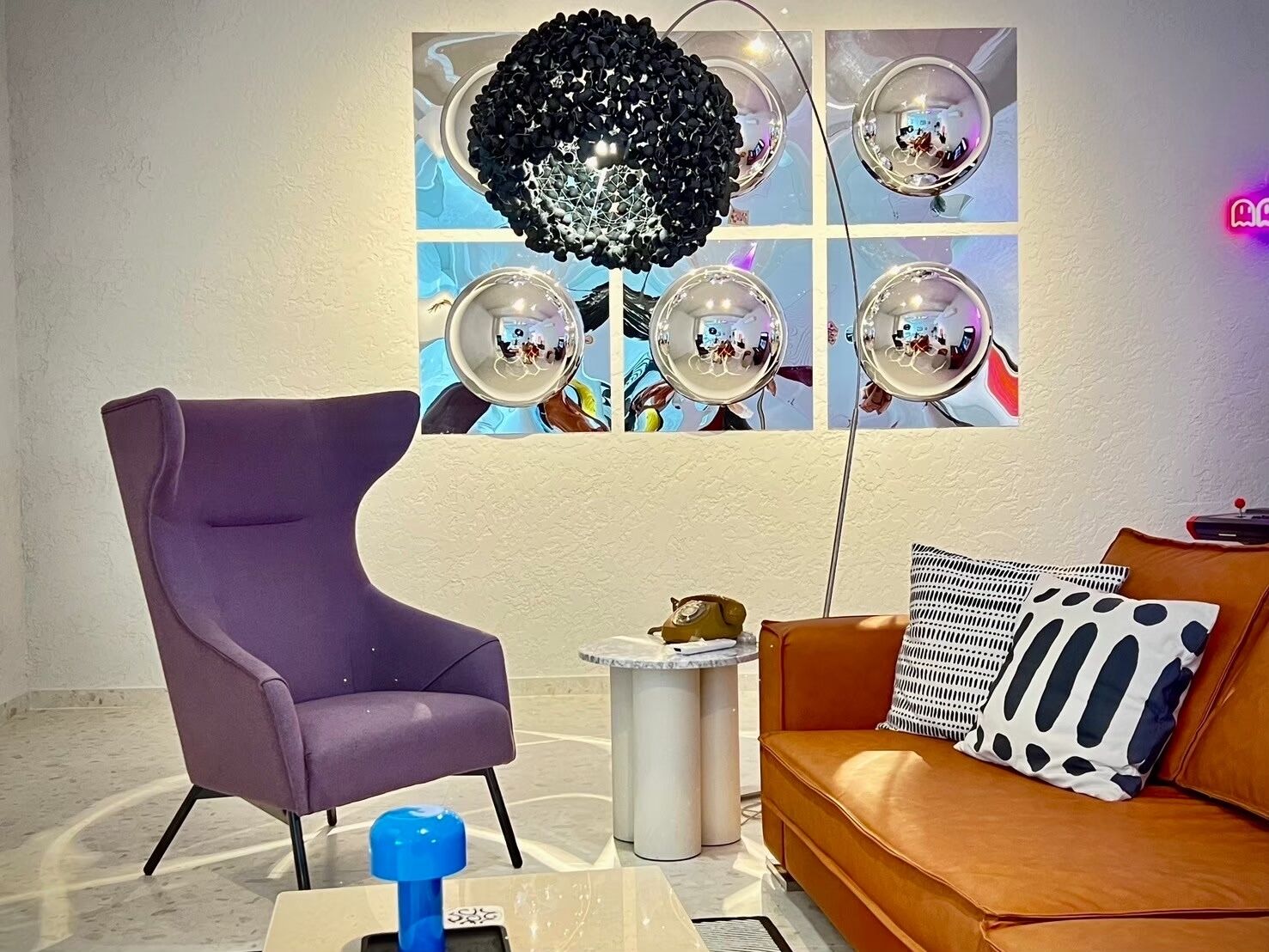 Uma sala de estar moderna com uma poltrona roxa, um sofá marrom com almofadas estampadas, uma mesinha redonda e uma peça de arte original em formato de esfera na parede.