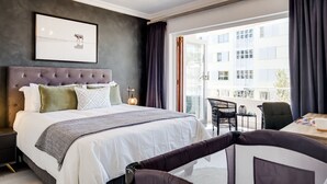 Quarto de hotel moderno com uma cama grande, cabeceira estofada, quadro emoldurado na parede, varanda com vista para edifícios e área de refeições com cadeiras e mesa.
