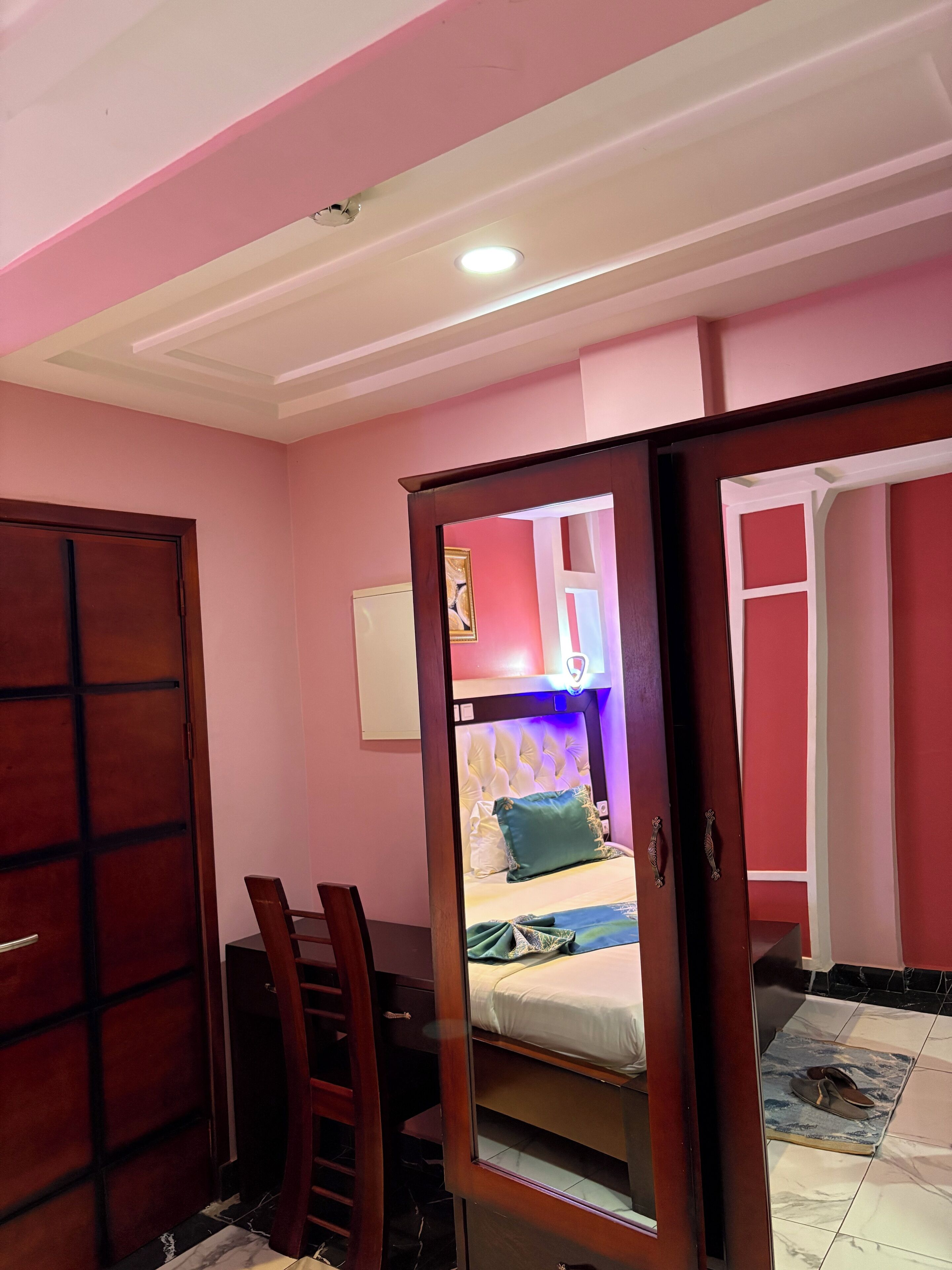 Quarto de hotel com paredes cor-de-rosa, um guarda-roupa de madeira, uma cama com cabeceira branca e uma pequena mesa com uma cadeira.