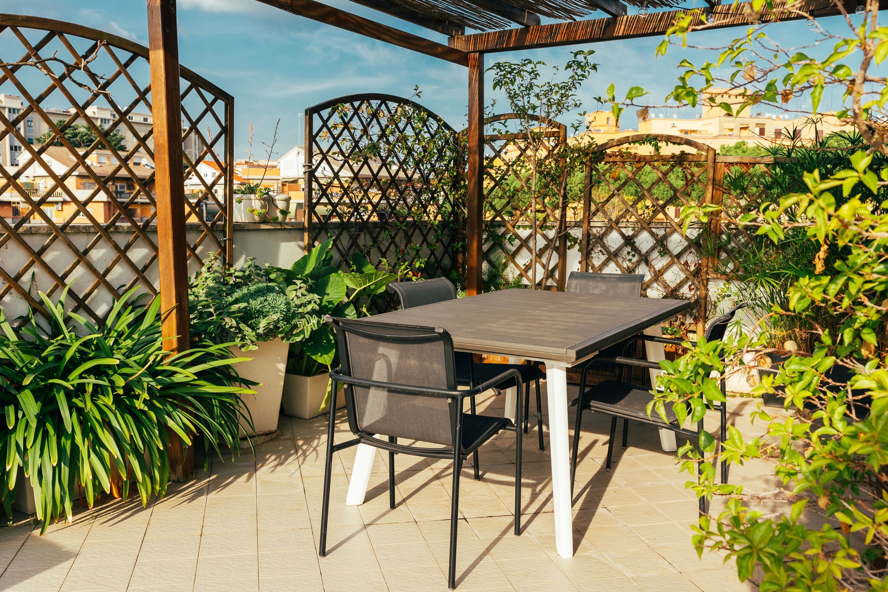 Um pátio externo com mesa e cadeiras de madeira, cercado por plantas em vasos e um cercado de treliça.